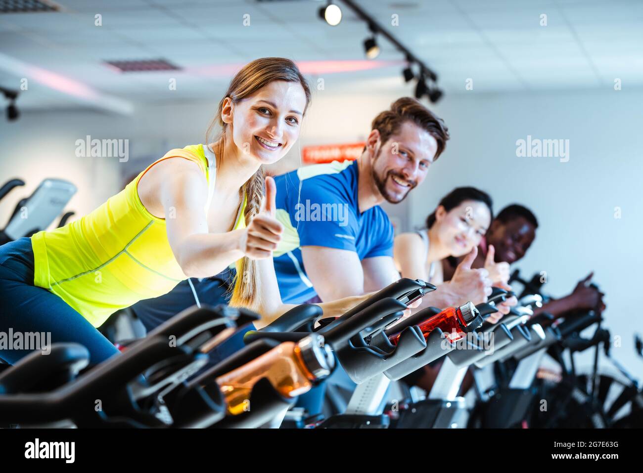 Woman showing thumbs up sign exercising of fitness bike in gym looking ...