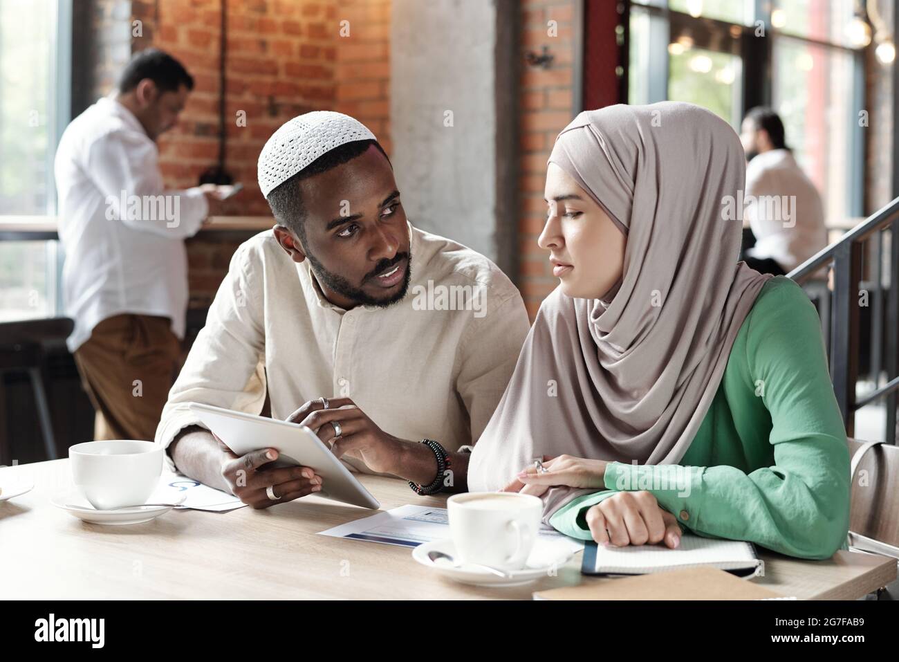 Young Black Man In Kufi Cap Using Tablet While Explaining Data To Female Colleague In Coffee