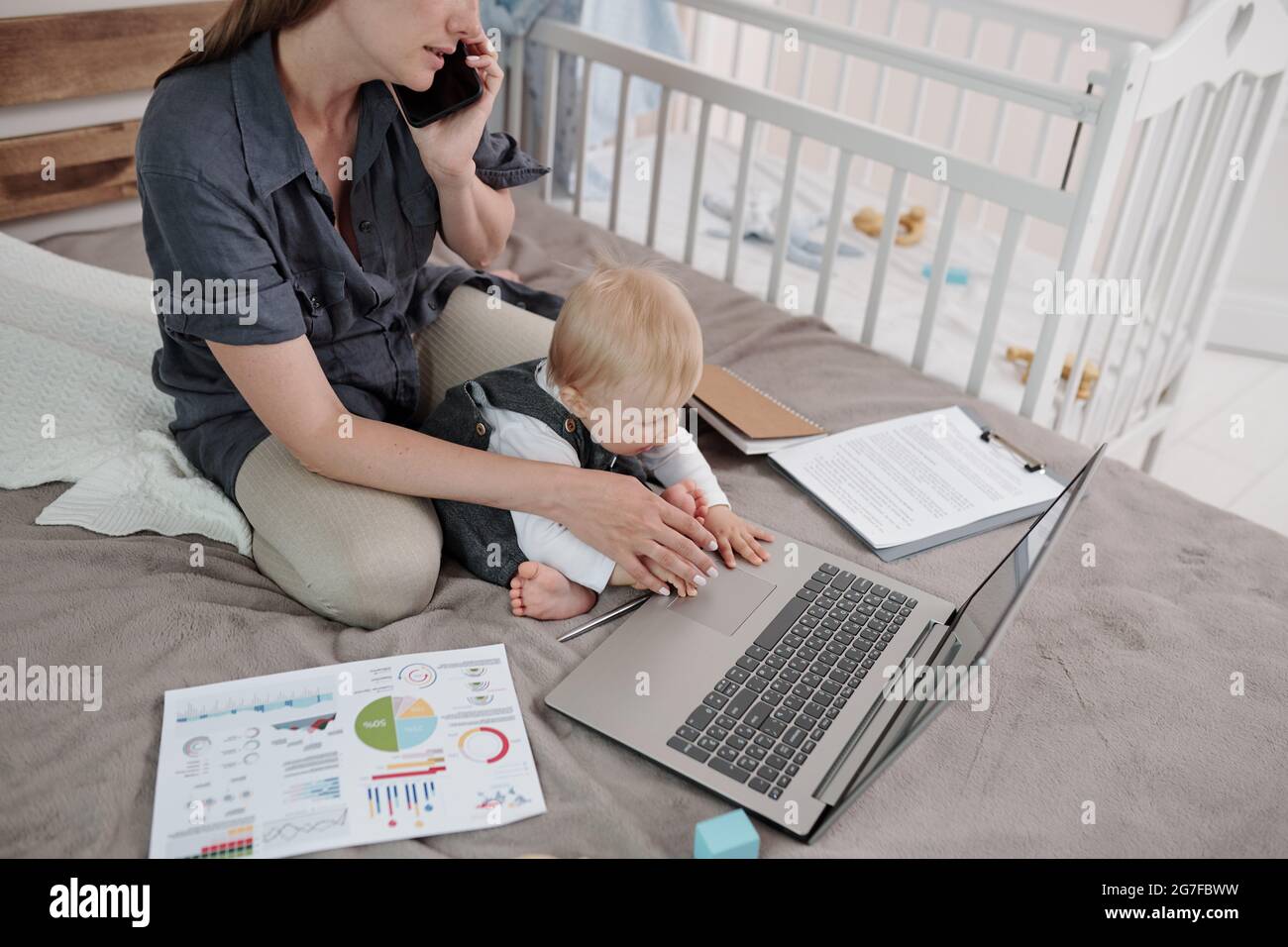 Multi-tasking young mother sitting with baby on bed and answering phone ...