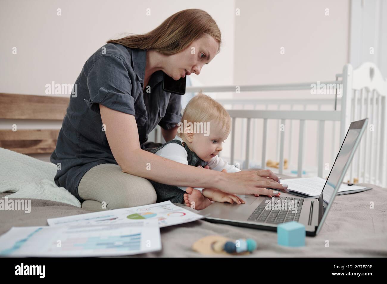 Serious young mother sitting on bed with papers and using laptop while ...