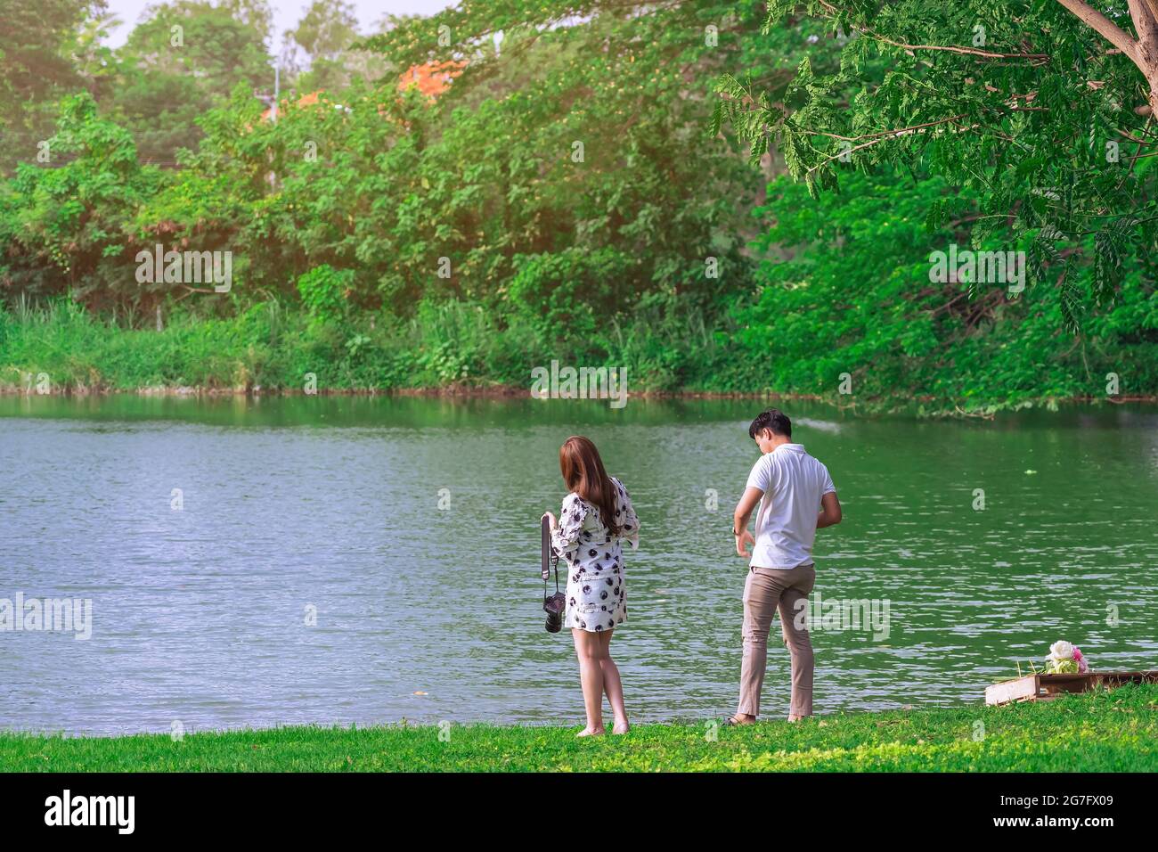 Back view of Asian lovers with camera relaxing near pond in garden ...