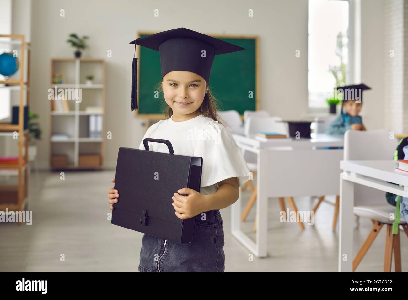 Portrait of happy pretty little student girl in academic cap standing ...