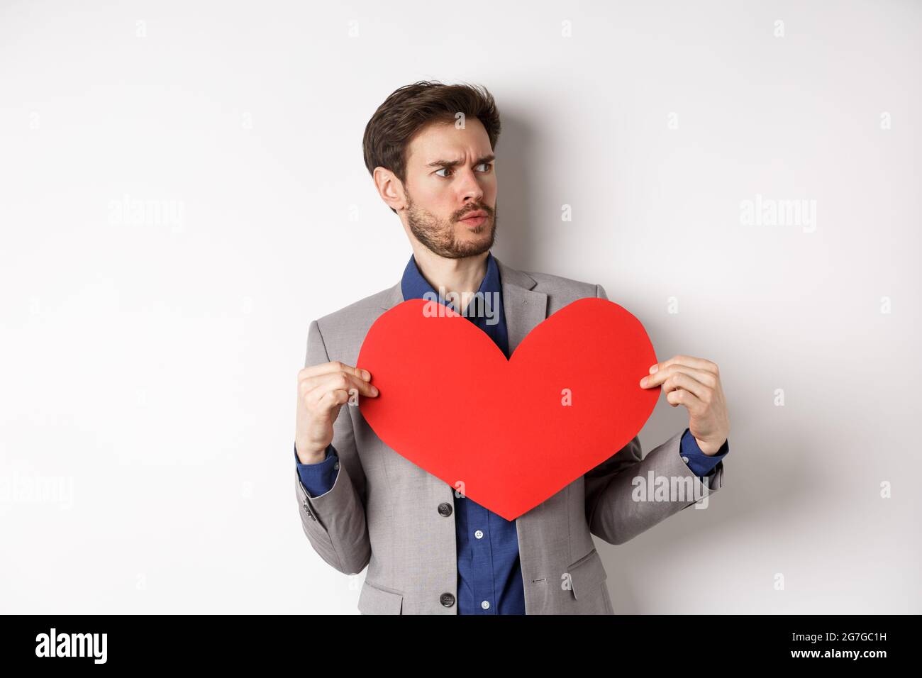 Confused man in suit holding big red heart and looking right, searching ...