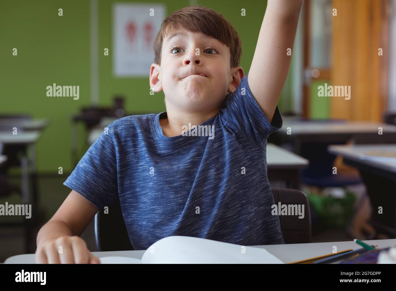 Excited caucasian schoolboy in classroom sitting at desk and raising ...