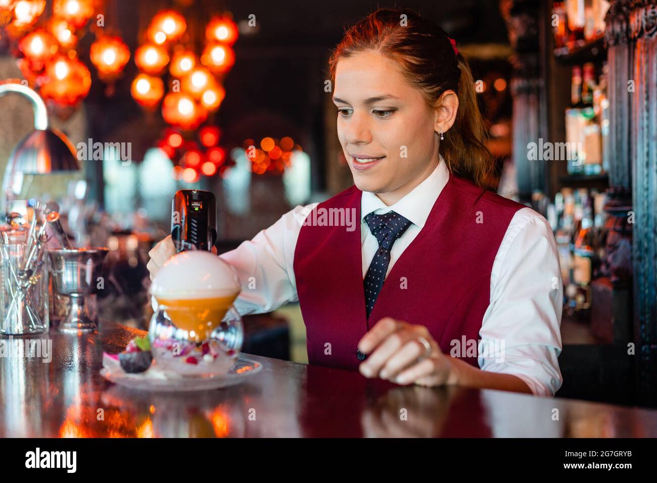 Skilled young female barkeeper using flavor bluster smoke gun while ...
