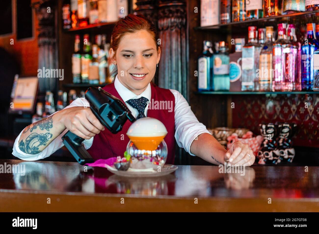 Skilled young female barkeeper using flavor bluster smoke gun while ...