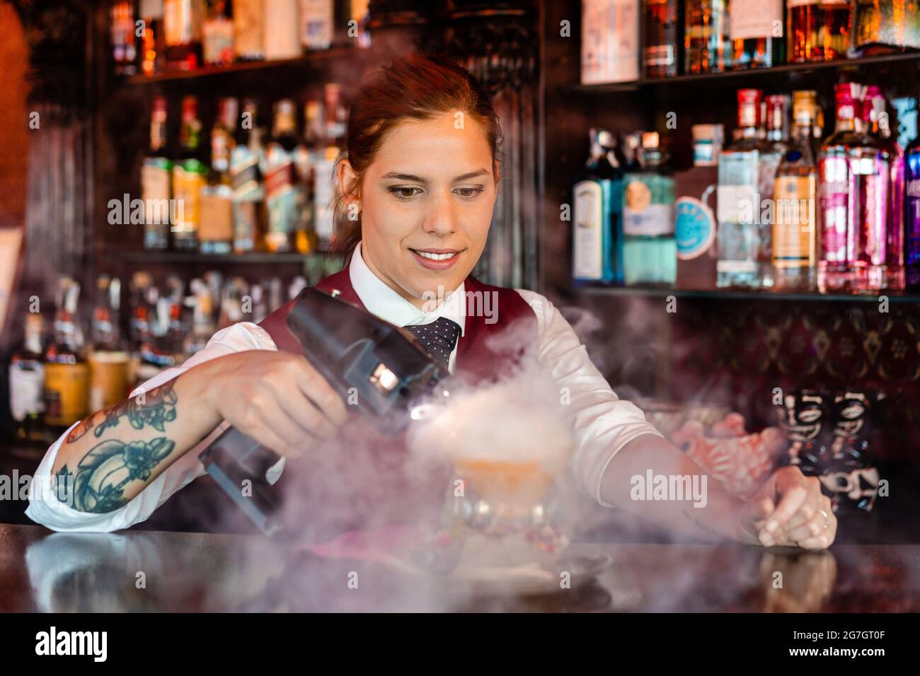 Skilled young female barkeeper using flavor bluster smoke gun while ...