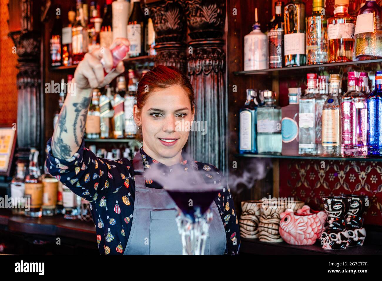 Delighted female barkeeper in apron preparing flavor blaster cocktail ...