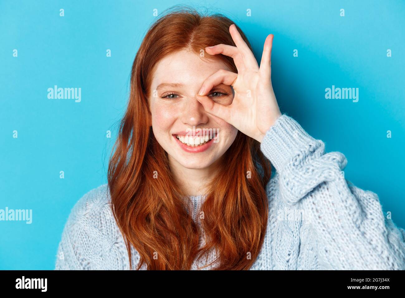 Headshot of cheerful redhead female model showing okay sign over eye ...