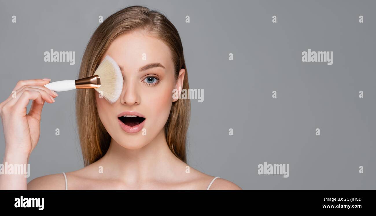 amazed young woman holding cosmetic brush isolated on grey, banner ...