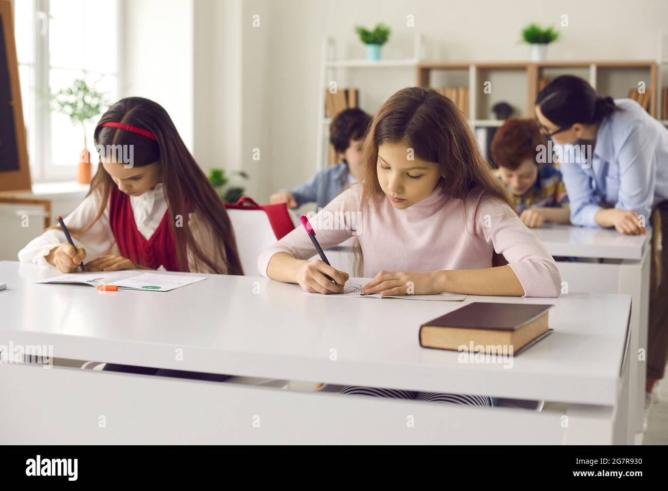 Smart elementary pupil writing in notebook sitting at desk in classroom ...