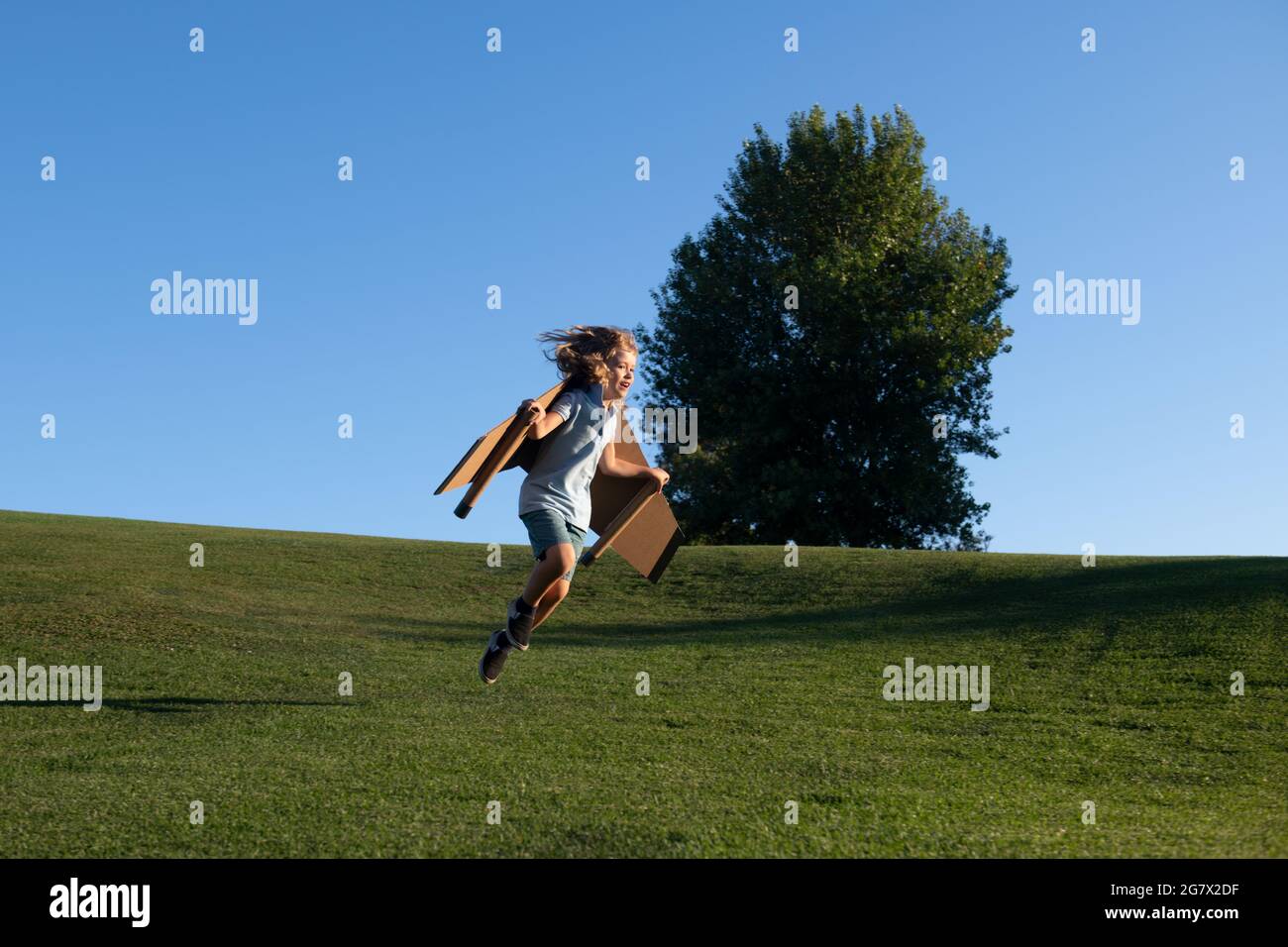 Kid running jumping and flying with toy plane wings on grass in park ...