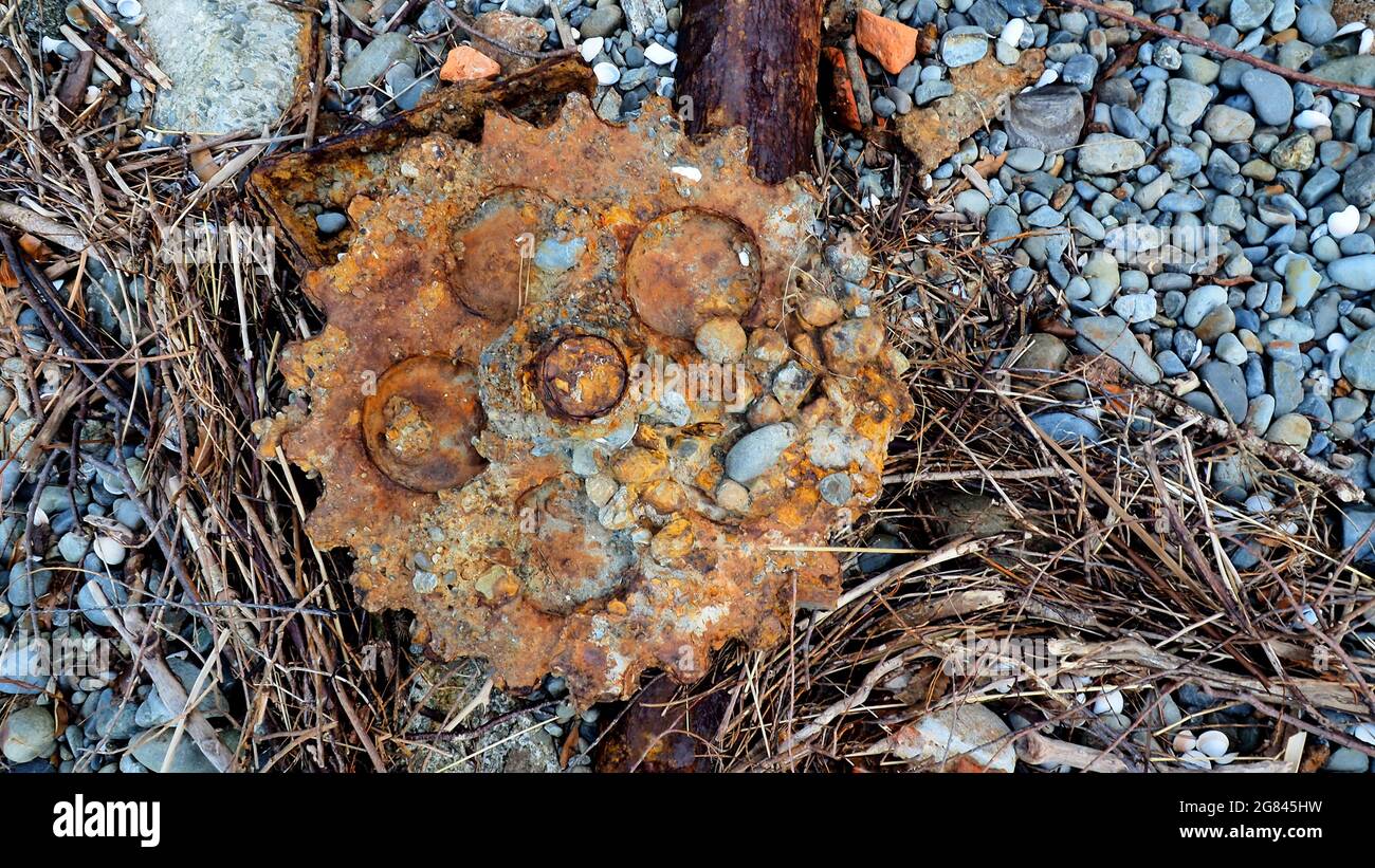 A rusted mechanical gear lies dumped on a river shore with stones and ...