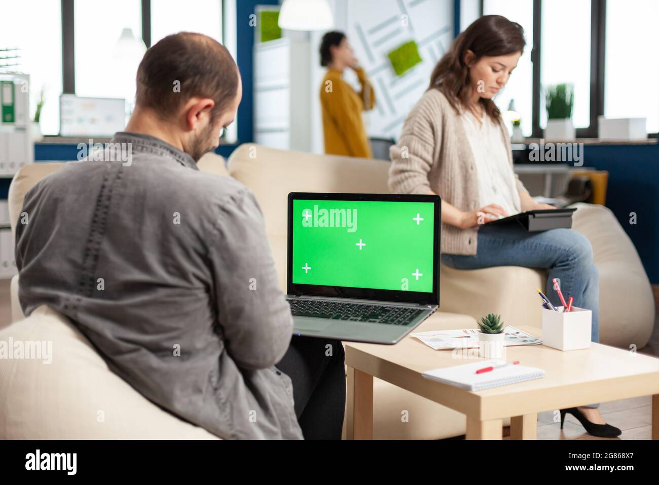 Over the sholder of manager sitting on couch holding laptop with green ...