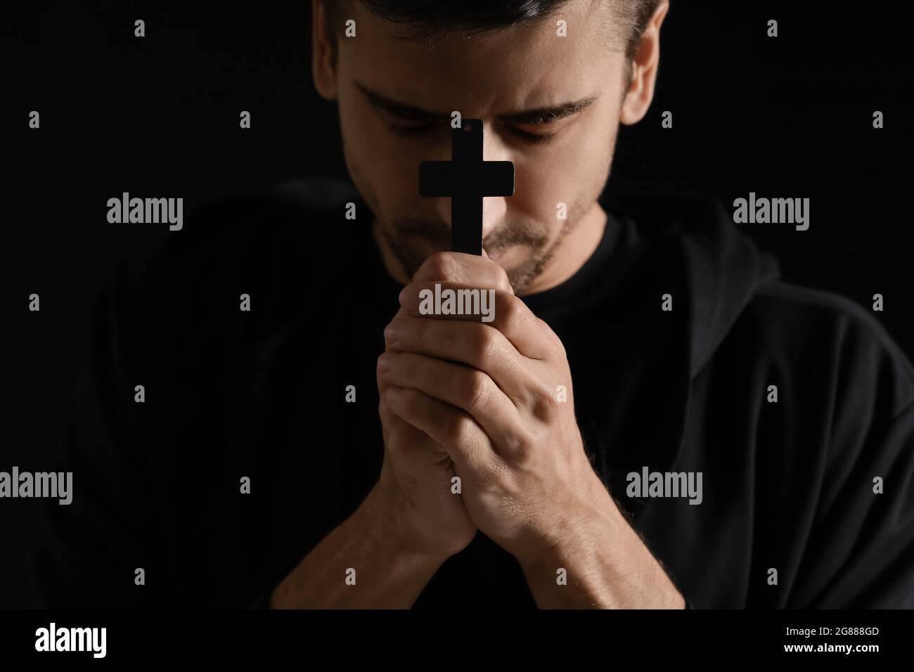Religious young man with cross praying on dark background, closeup ...