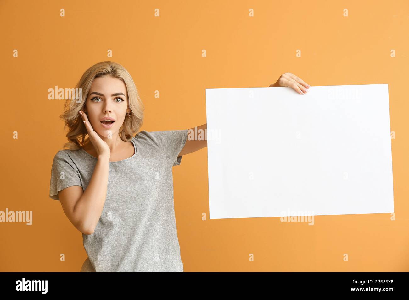 Surprised young woman with blank poster on color background Stock Photo ...