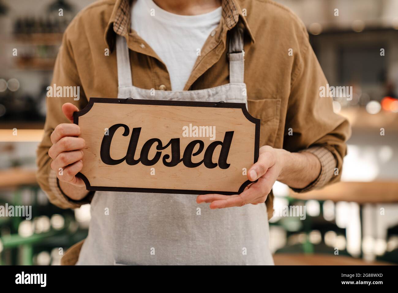 Young man waiter in apron showing closed sign while standing in cafe ...