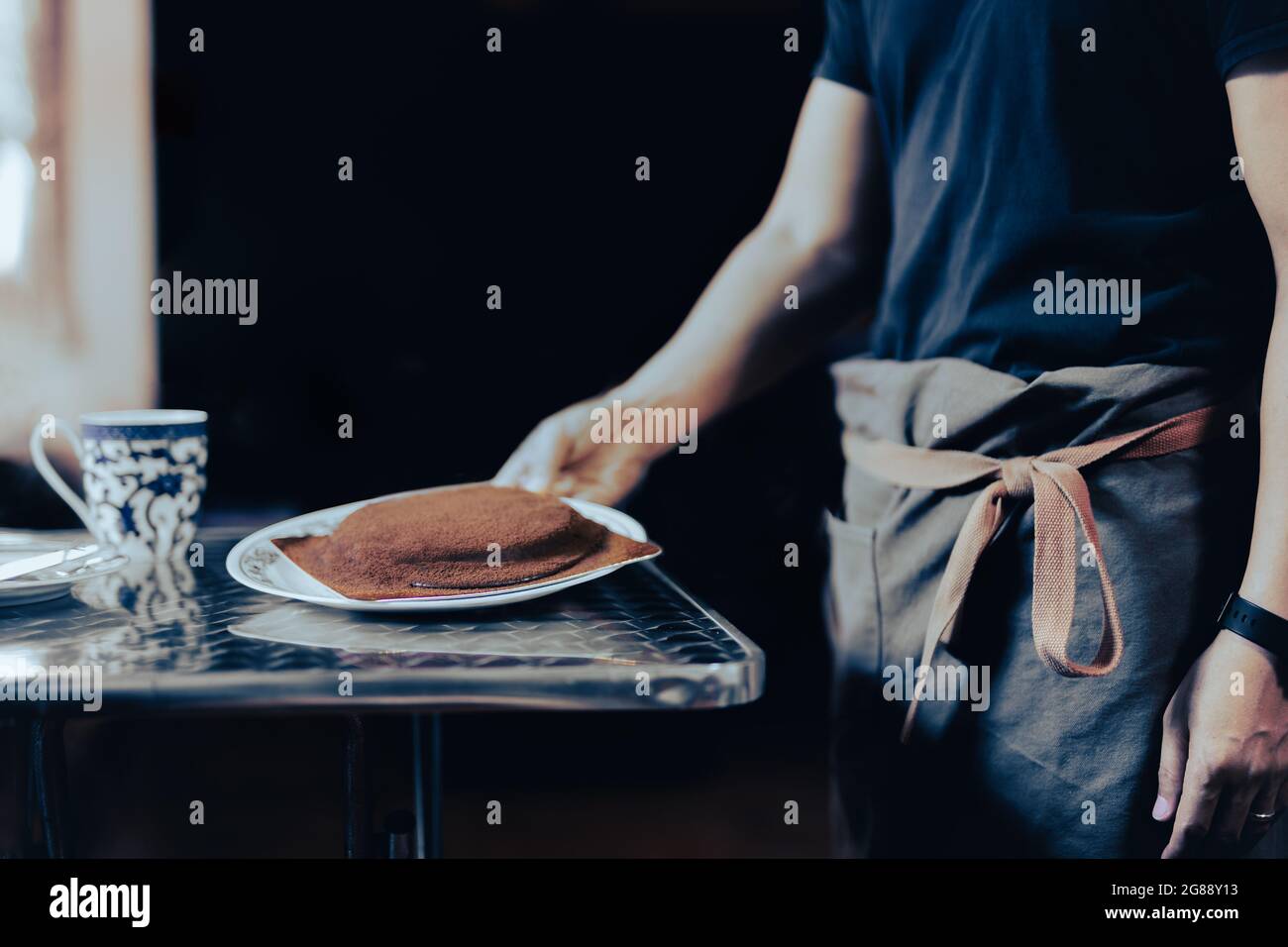 Waiter in apron serving whole chocolate cake at cafe in dark background ...