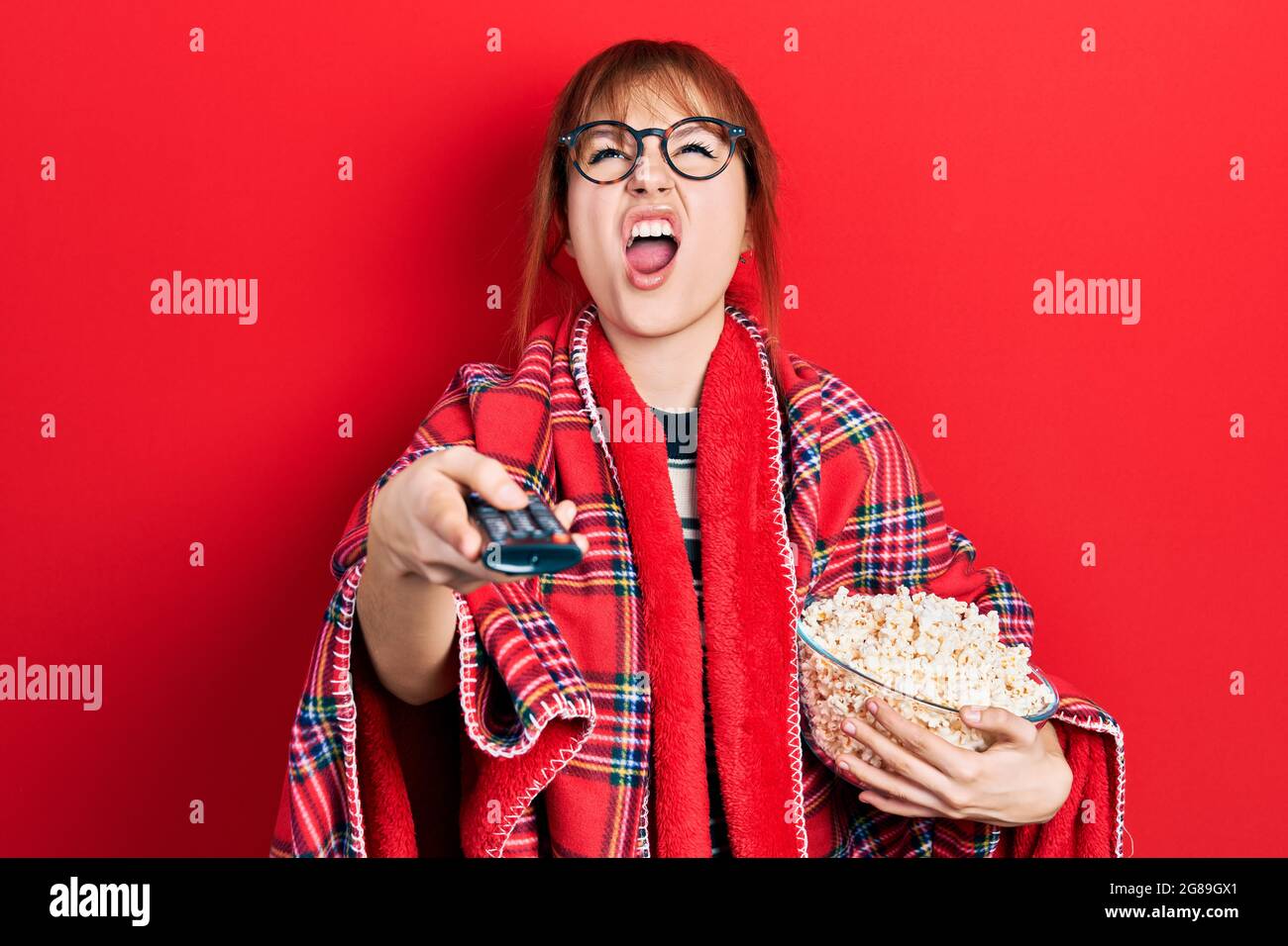 Redhead young woman eating popcorn using tv control angry and mad ...