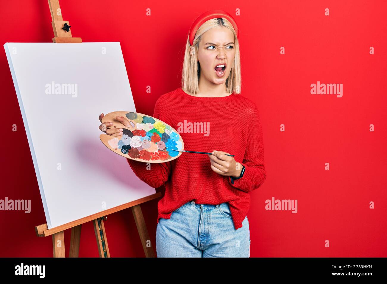 Beautiful blonde woman standing drawing with palette by painter easel ...