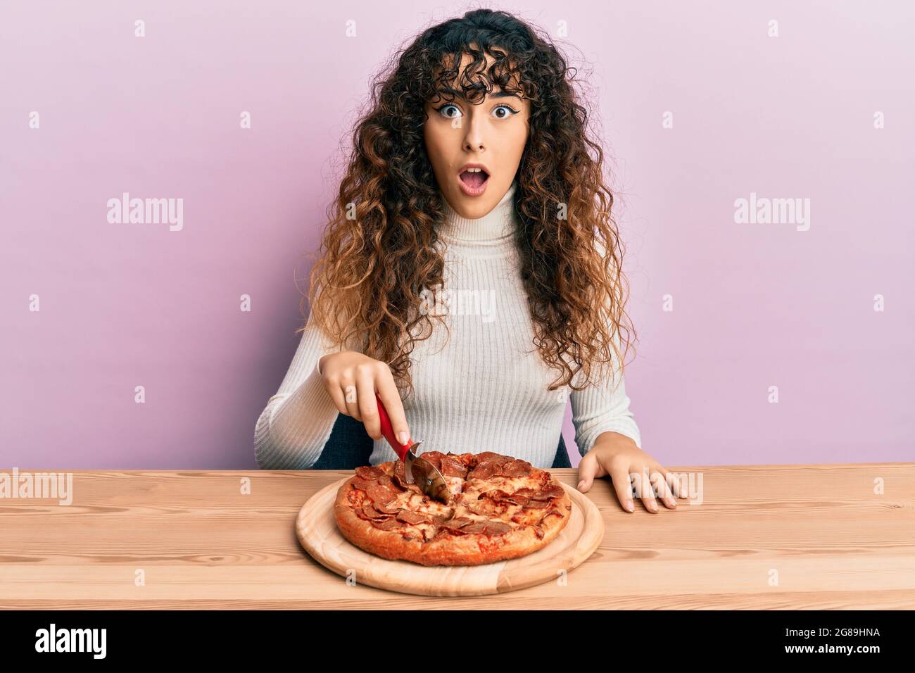 Young hispanic girl cutting tasty pepperoni pizza scared and amazed ...