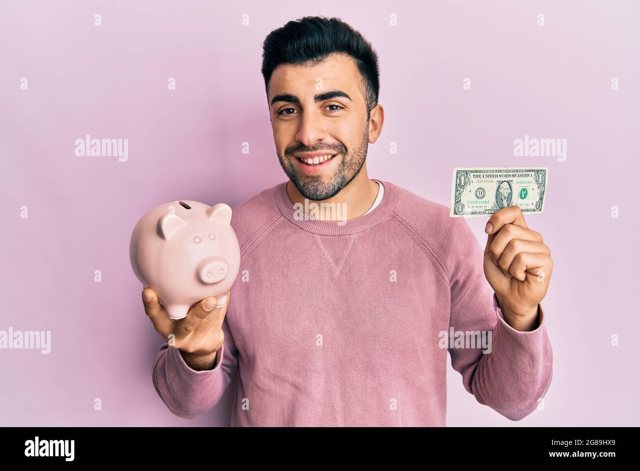 Young hispanic man holding one dollar banknote and piggy bank smiling ...