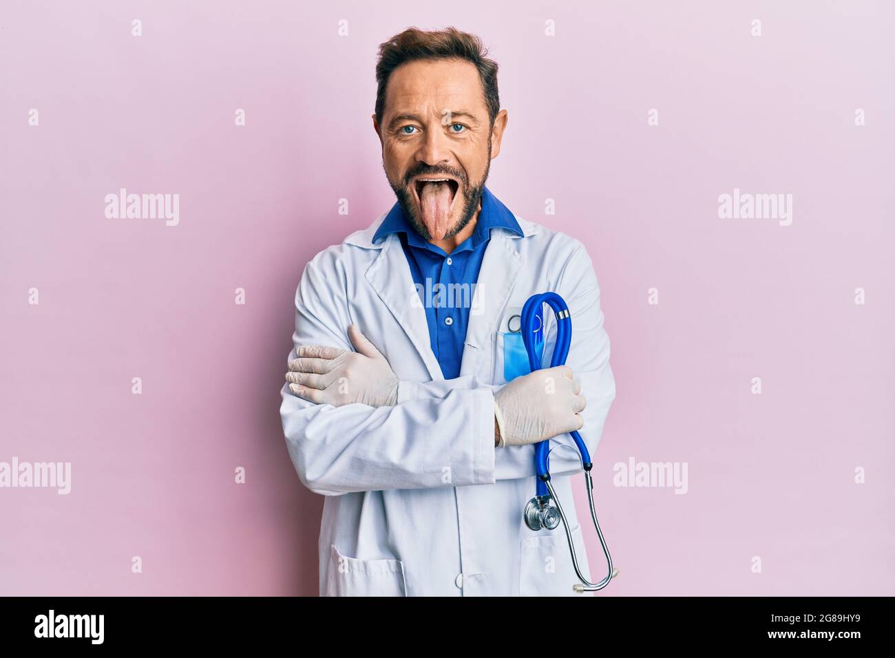 Middle age man wearing doctor uniform sticking tongue out happy with ...