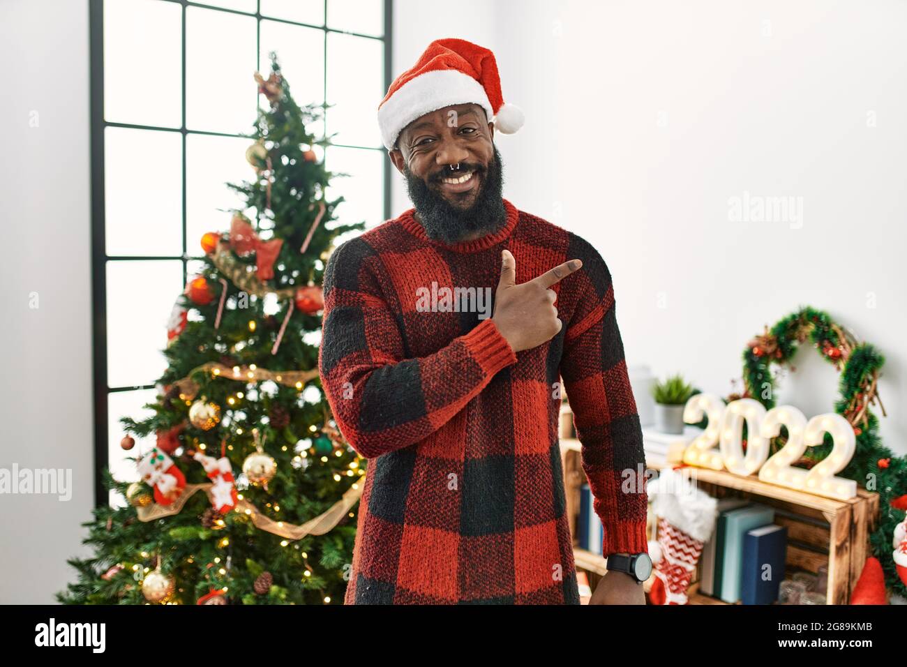 African american man wearing santa claus hat standing by christmas tree ...