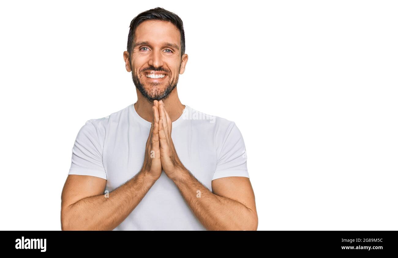 Handsome man with beard wearing casual white t shirt praying with hands ...