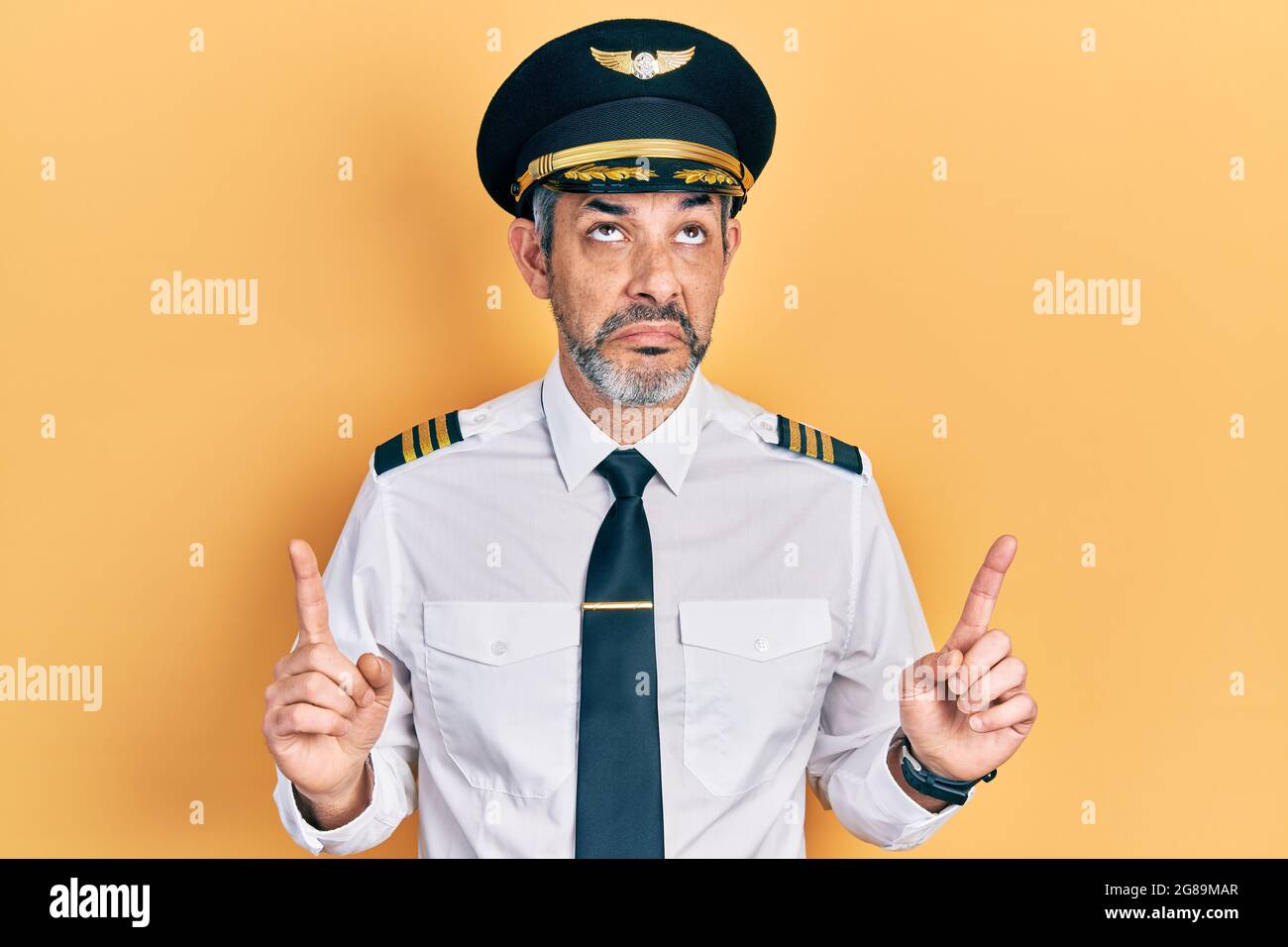 Handsome middle age man with grey hair wearing airplane pilot uniform pointing up looking sad ...