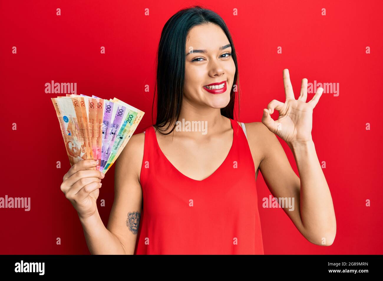 Young hispanic girl holding philippine peso banknotes doing ok sign ...