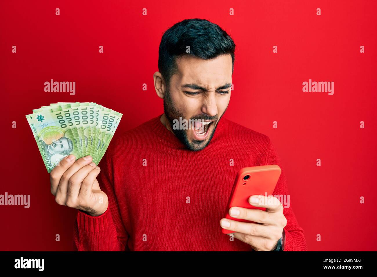 Young hispanic man using smartphone holding chilean pesos banknotes ...