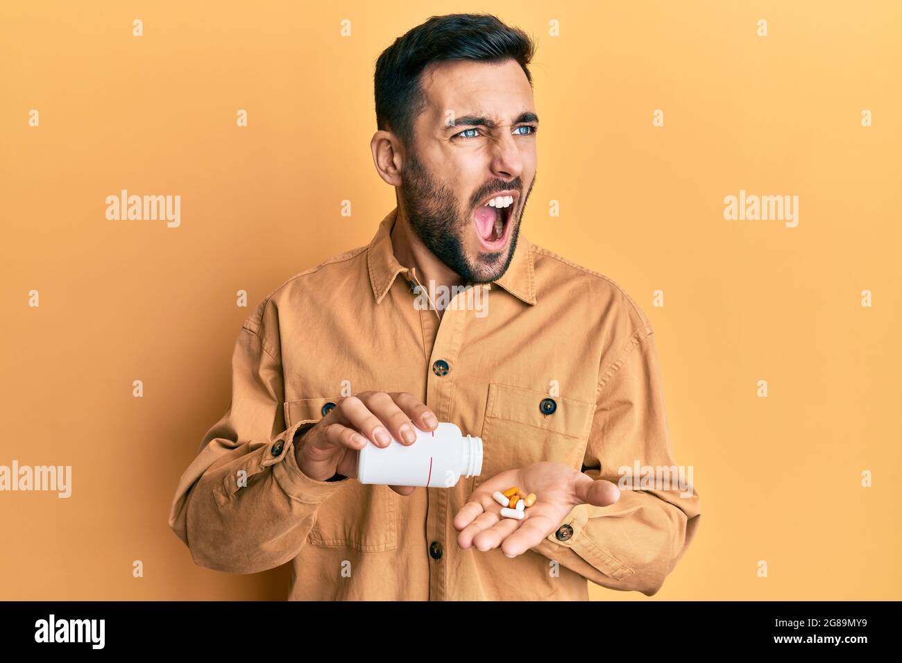 Young hispanic man holding pills angry and mad screaming frustrated and ...