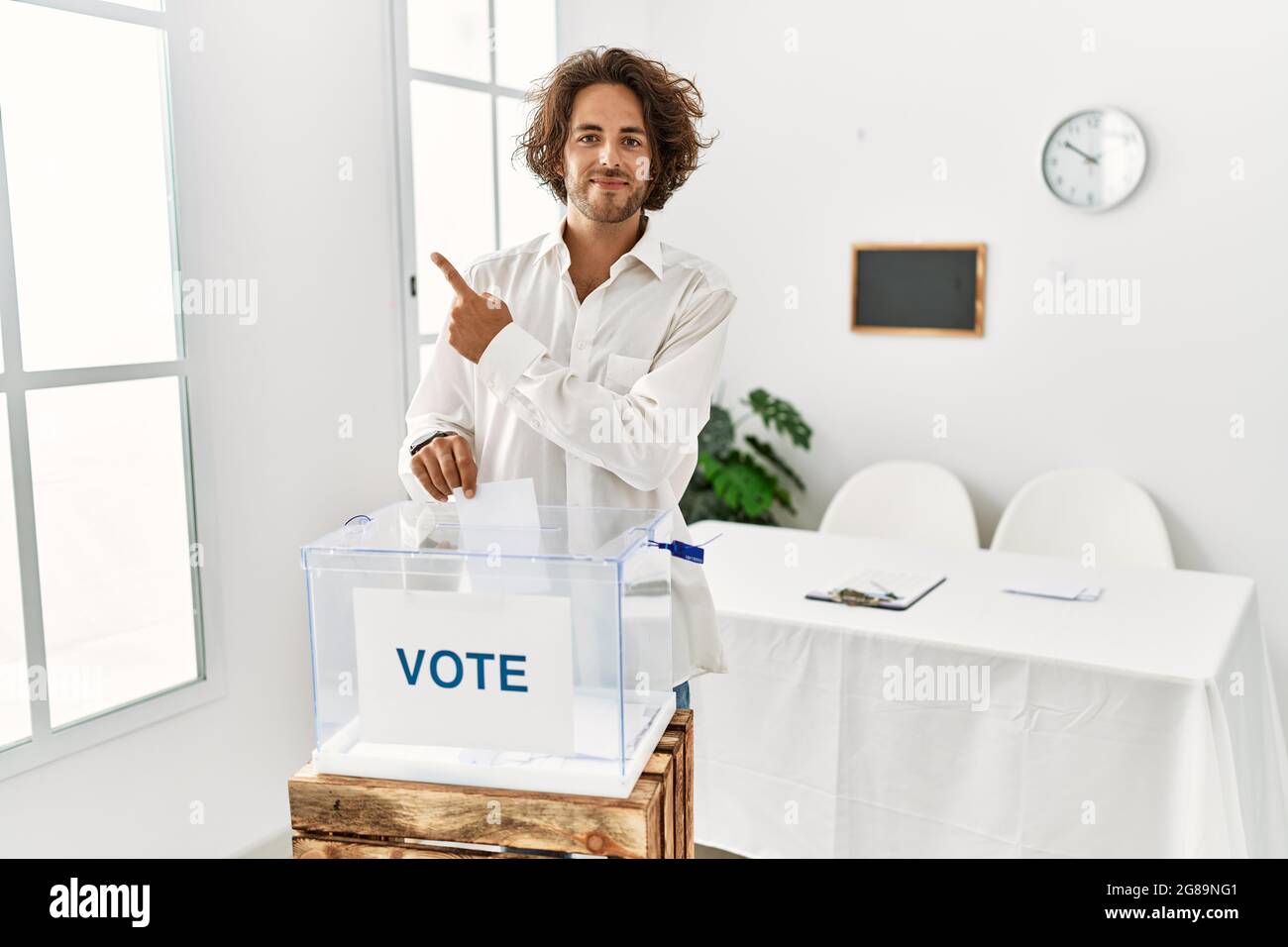 Young hispanic man voting putting envelop in ballot box pointing with ...