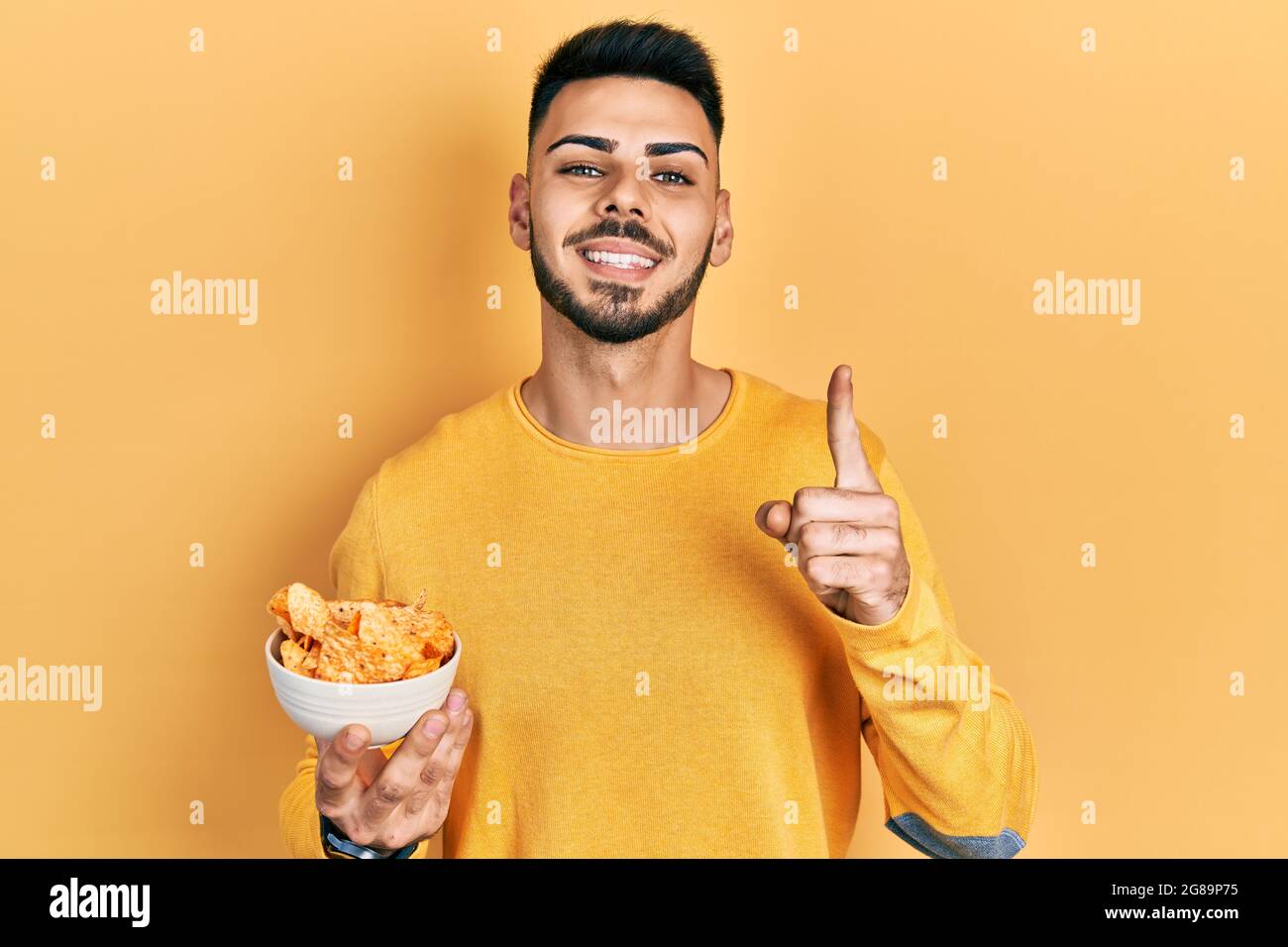 Young hispanic man with beard holding nachos potato chips smiling with ...