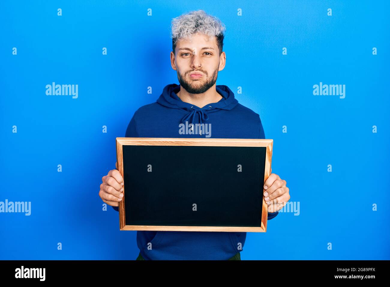 Young hispanic man with modern dyed hair holding blackboard puffing ...