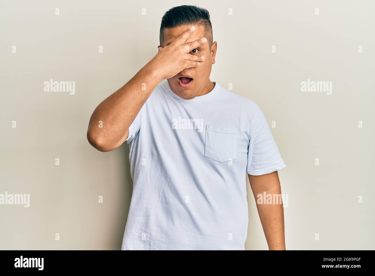 Young latin man wearing casual white t shirt peeking in shock covering ...