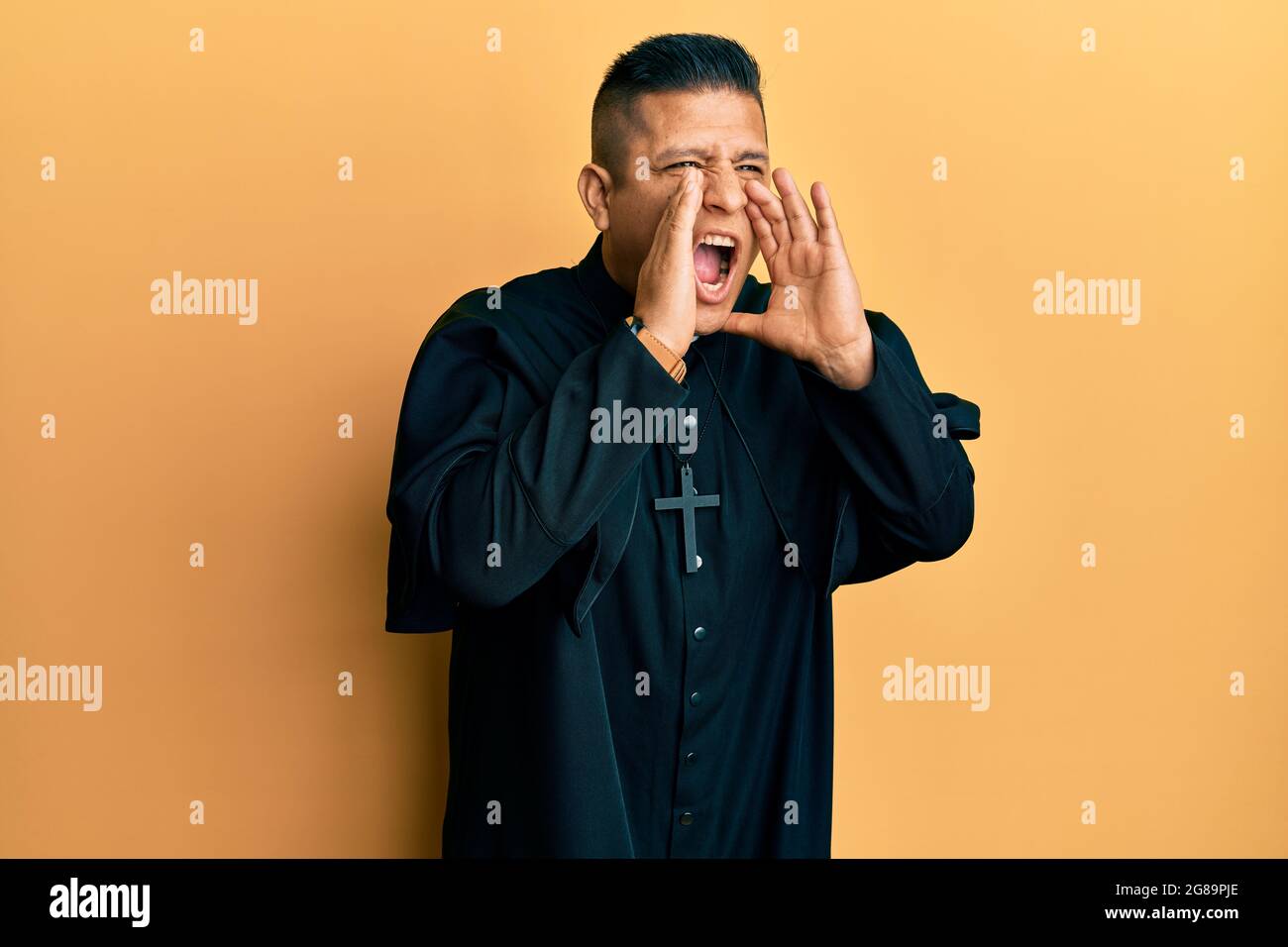 Young latin priest man standing over yellow background shouting angry ...