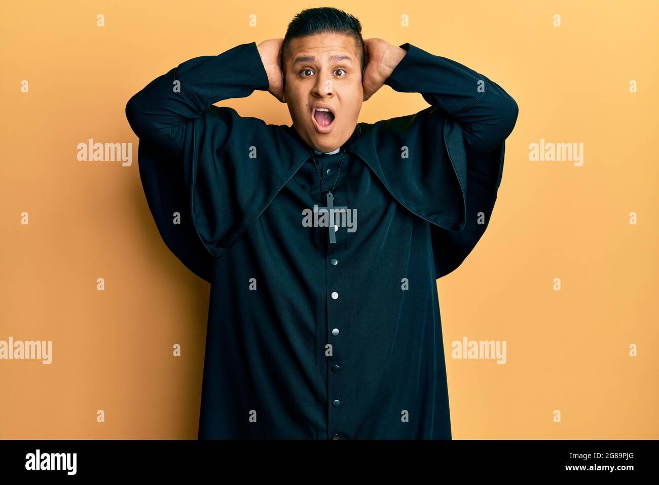 Young latin priest man standing over yellow background crazy and scared ...