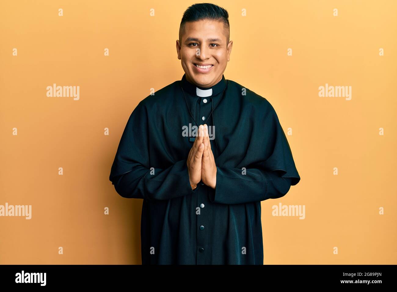Young latin priest man praying with hands together smiling with a happy ...