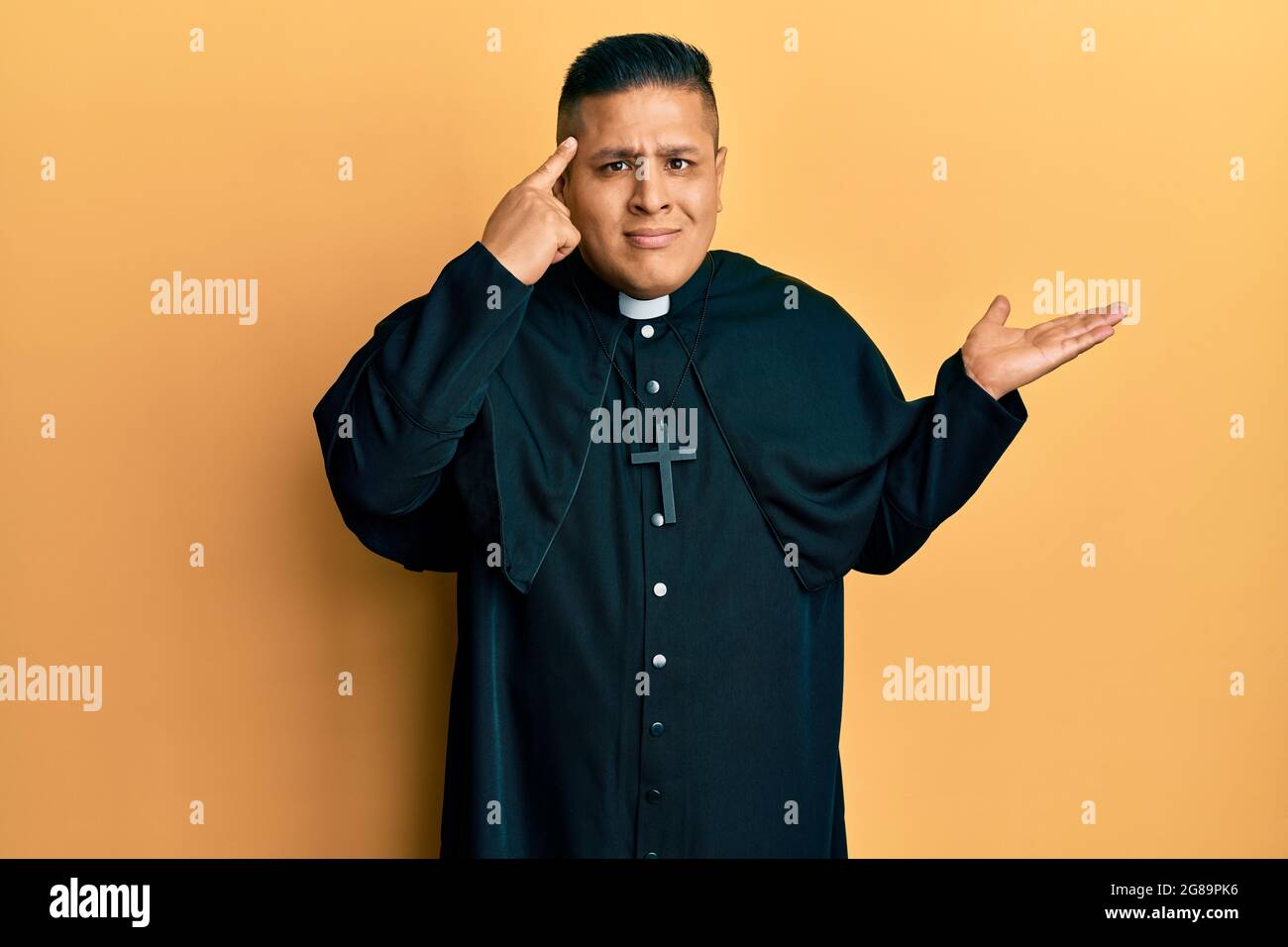 Young latin priest man standing over yellow background confused and ...