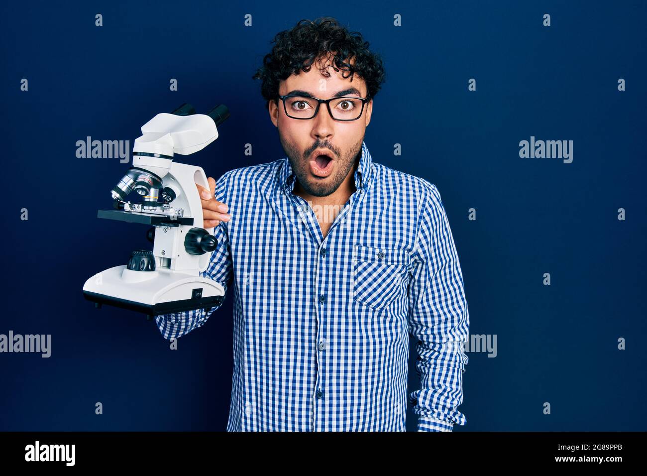 Young hispanic man holding microscope scared and amazed with open mouth ...