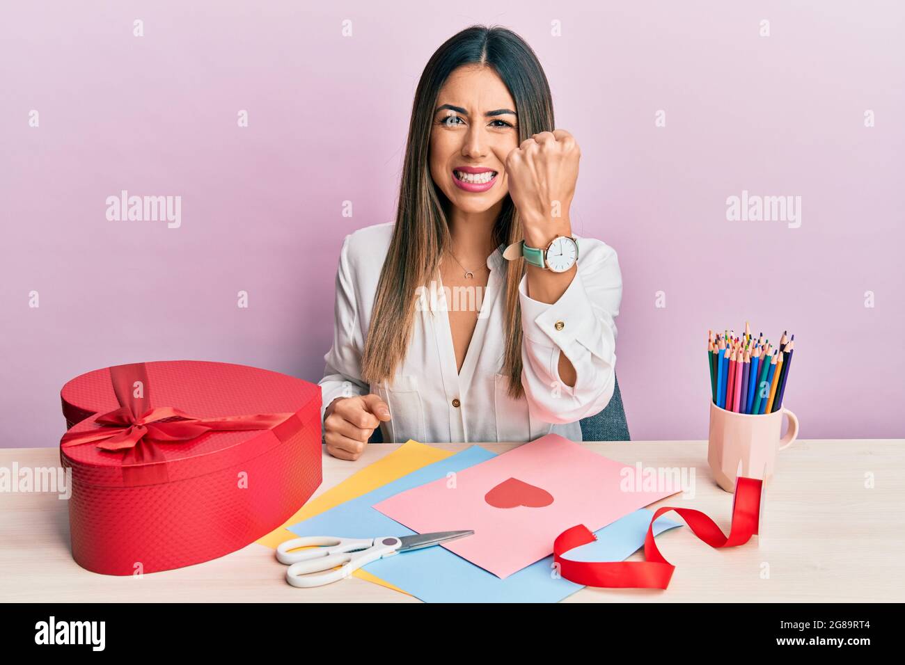 Young hispanic woman making valentine gift sitting on the table angry ...