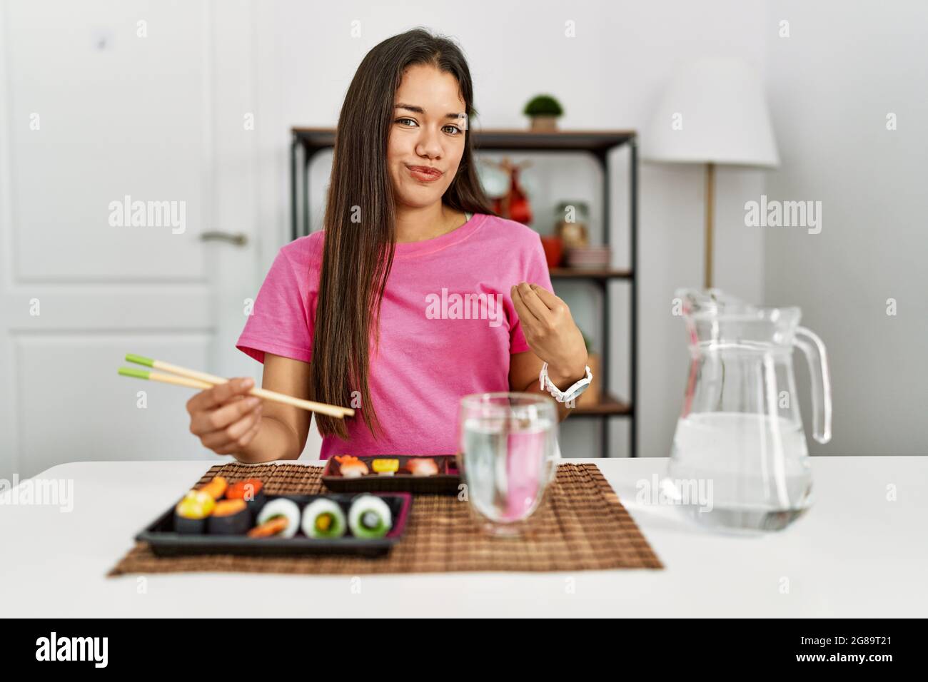 Young brunette woman eating sushi using chopsticks doing money gesture ...