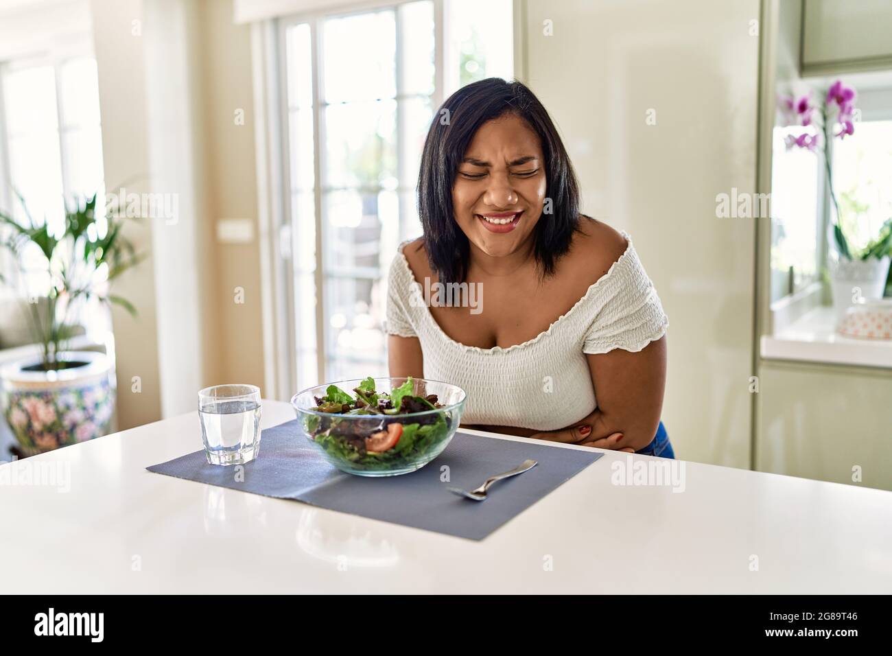 Young hispanic woman eating healthy salad at home with hand on stomach ...