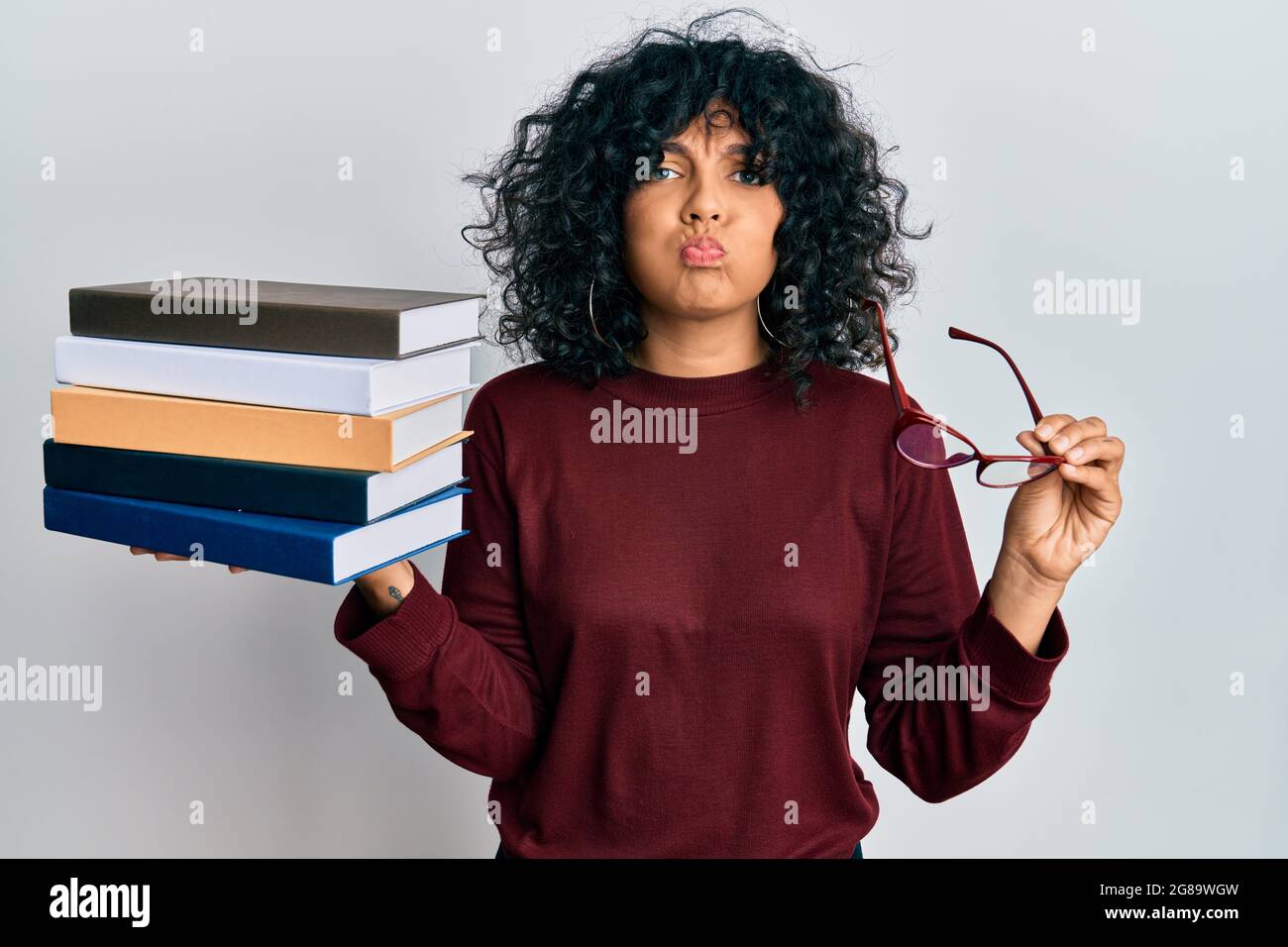 Young hispanic woman holding glasses and books puffing cheeks with ...