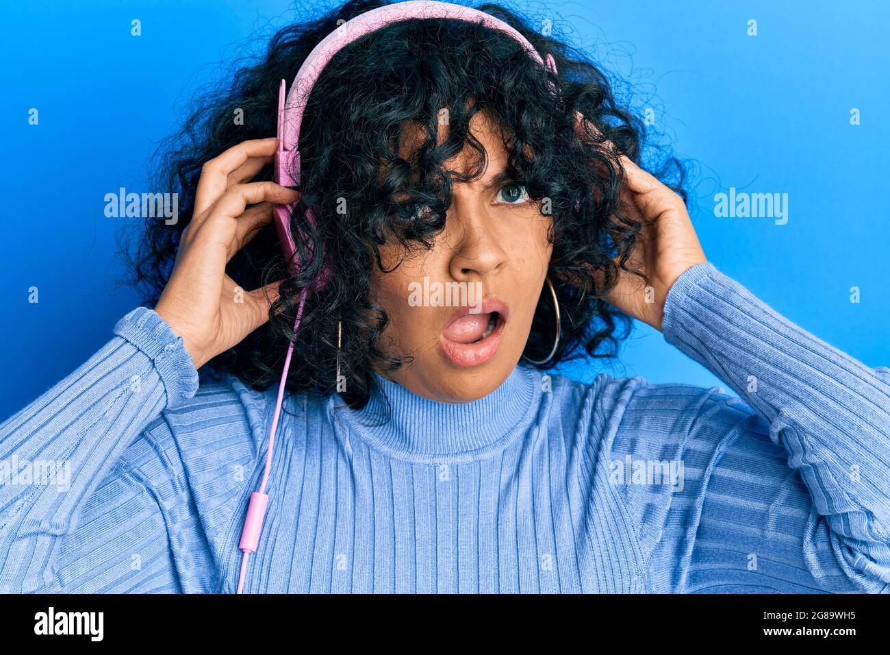 Young hispanic woman listening to music using headphones in shock face ...