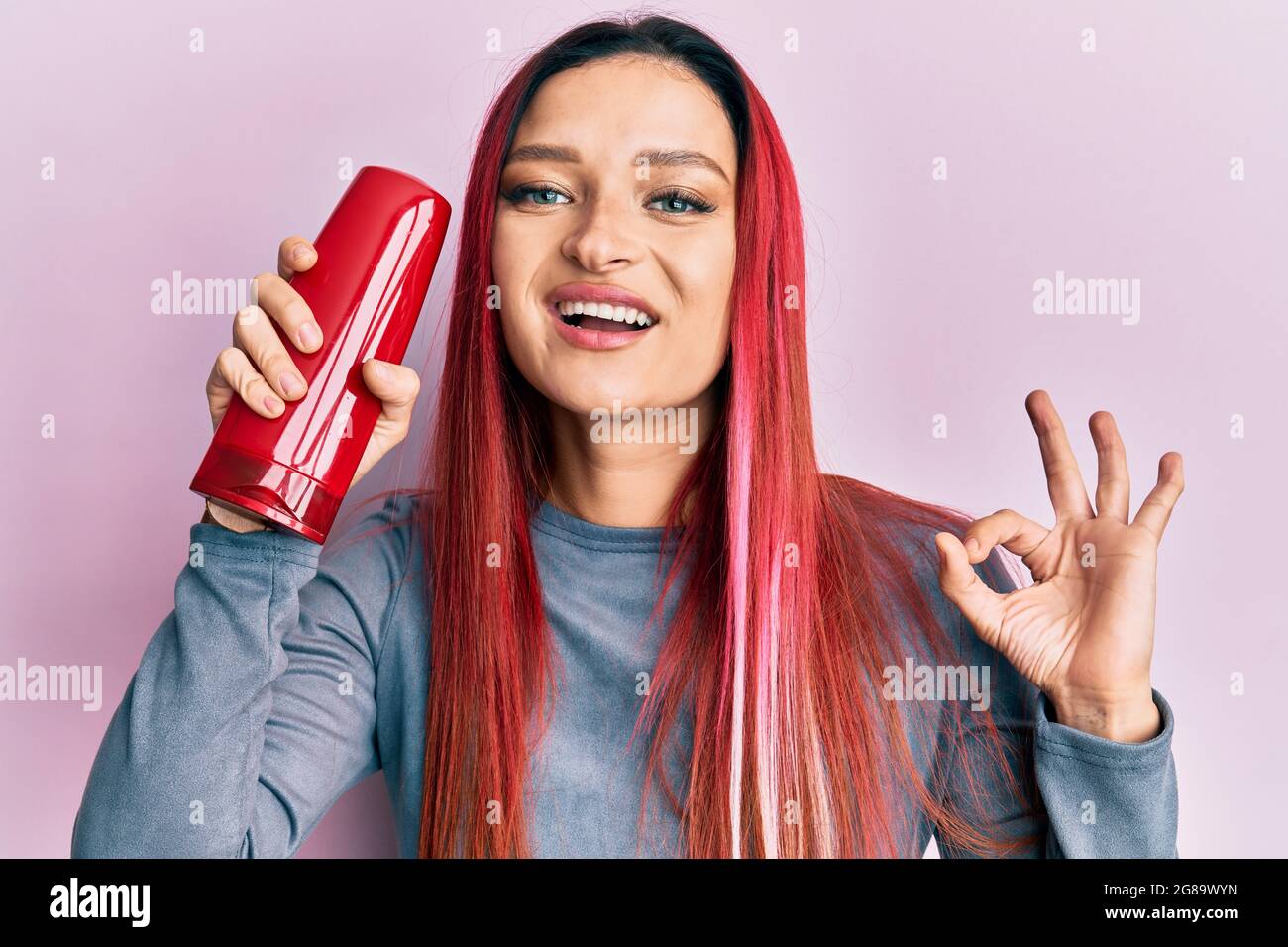 Young caucasian woman holding shampoo bottle doing ok sign with fingers ...