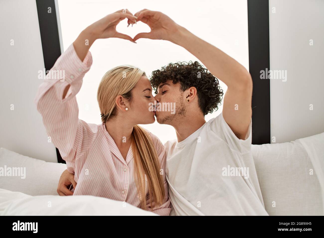 Young beautiful couple doing heart symbol with hands sitting on the bed ...
