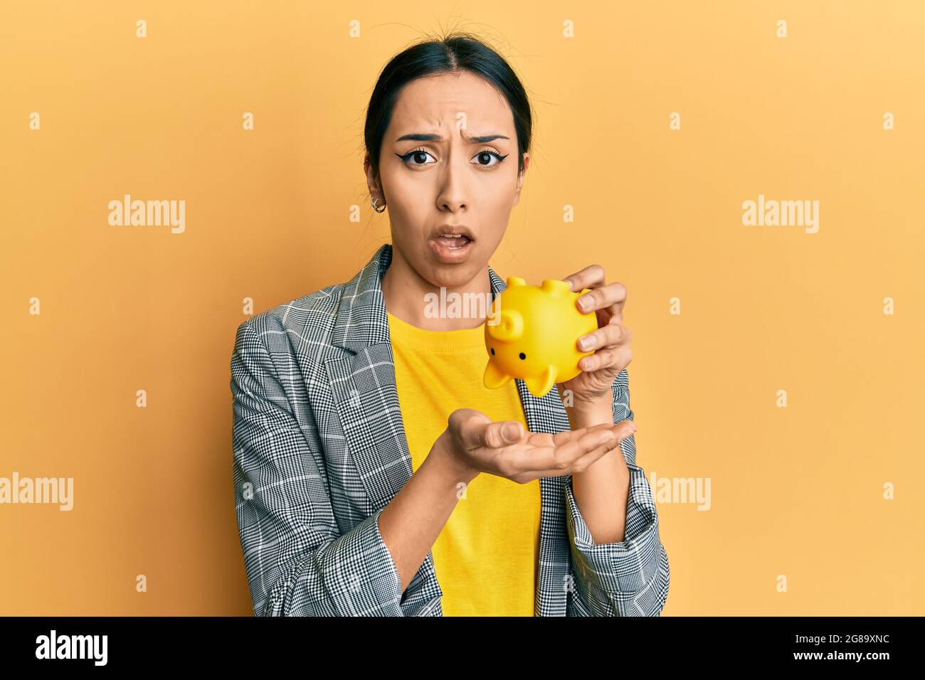 Young hispanic girl holding piggy bank in shock face, looking skeptical ...